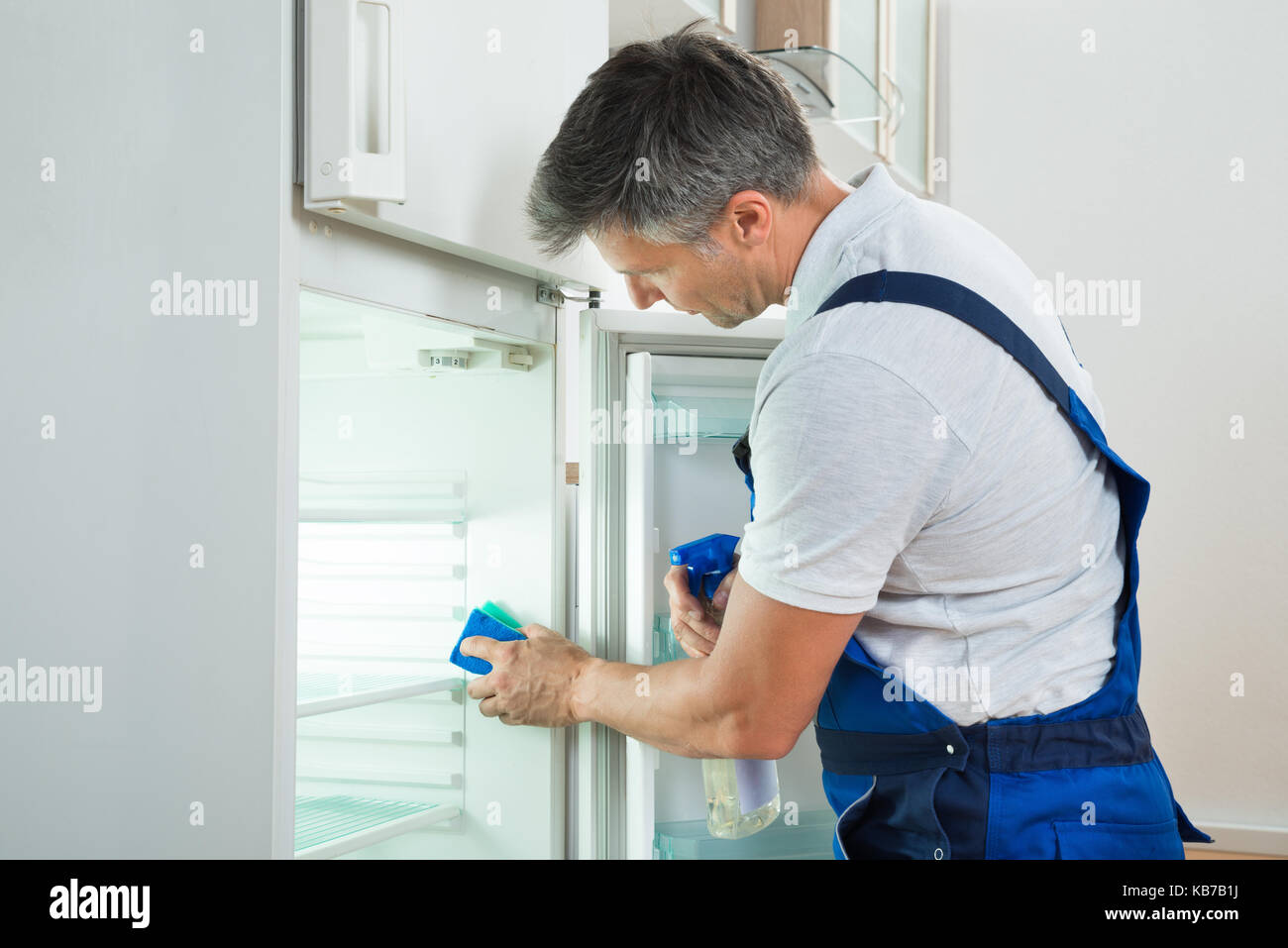 Side view of mature janitor cleaning refrigerator with spray bottle and ...