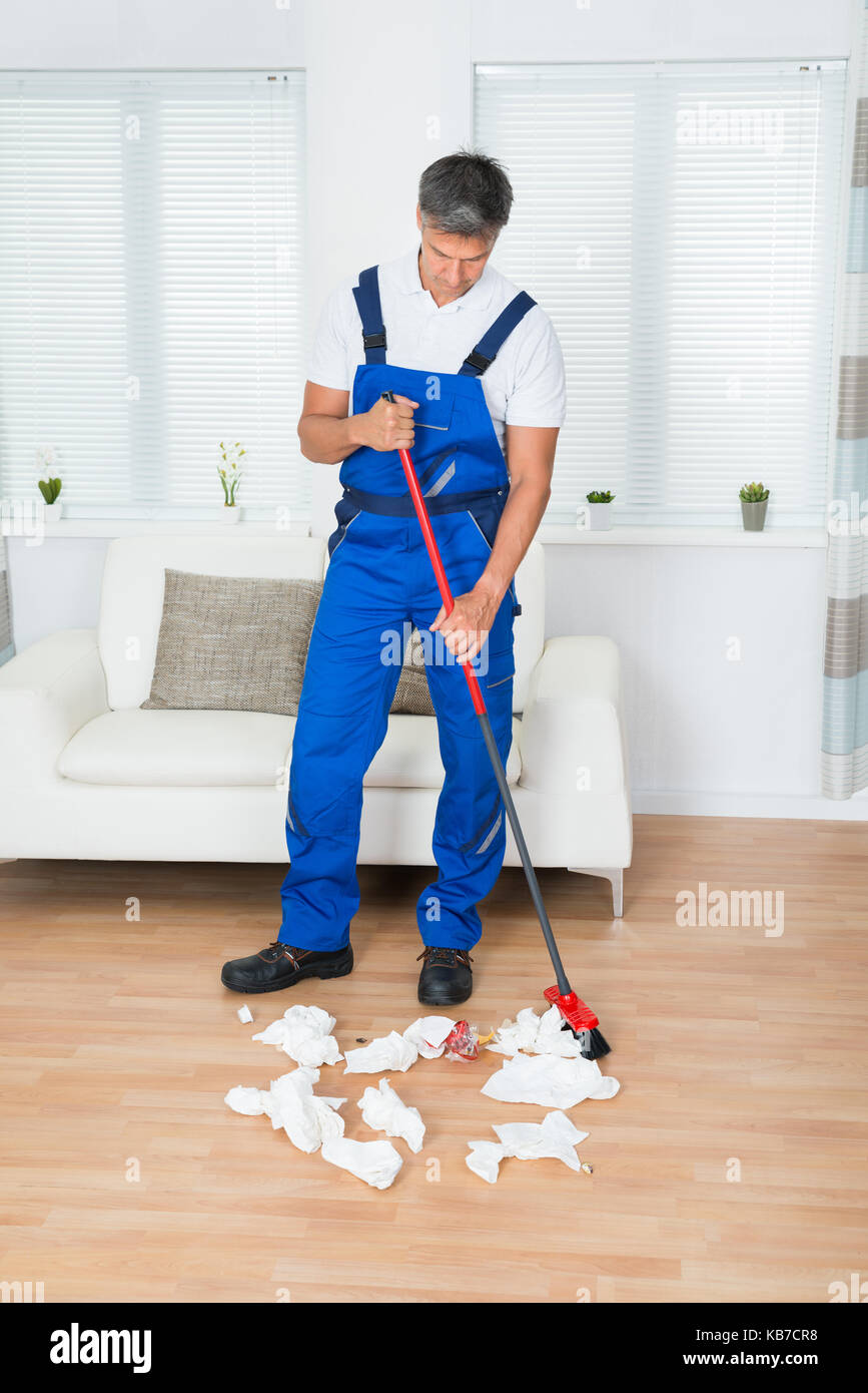 Full length of male janitor sweeping crumpled papers on floor in living ...