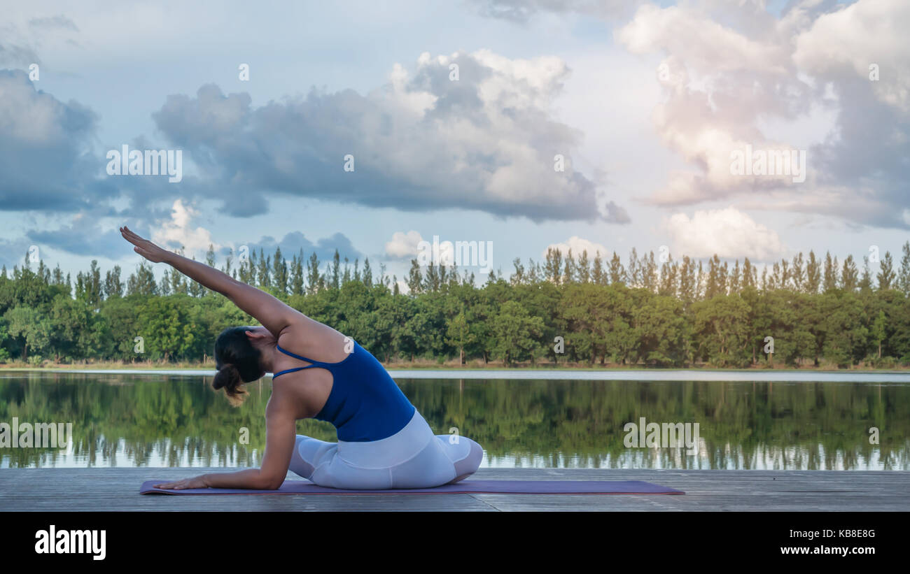 Asian woman practicing yoga pose , exercise outdoors with view of beautiful lake - relax in ...