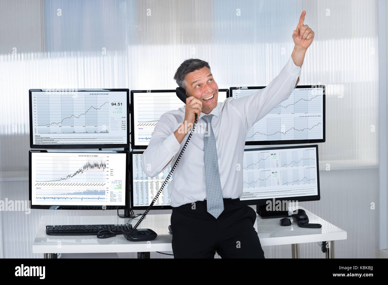 Happy male stock trader pointing upwards while using telephone at desk ...