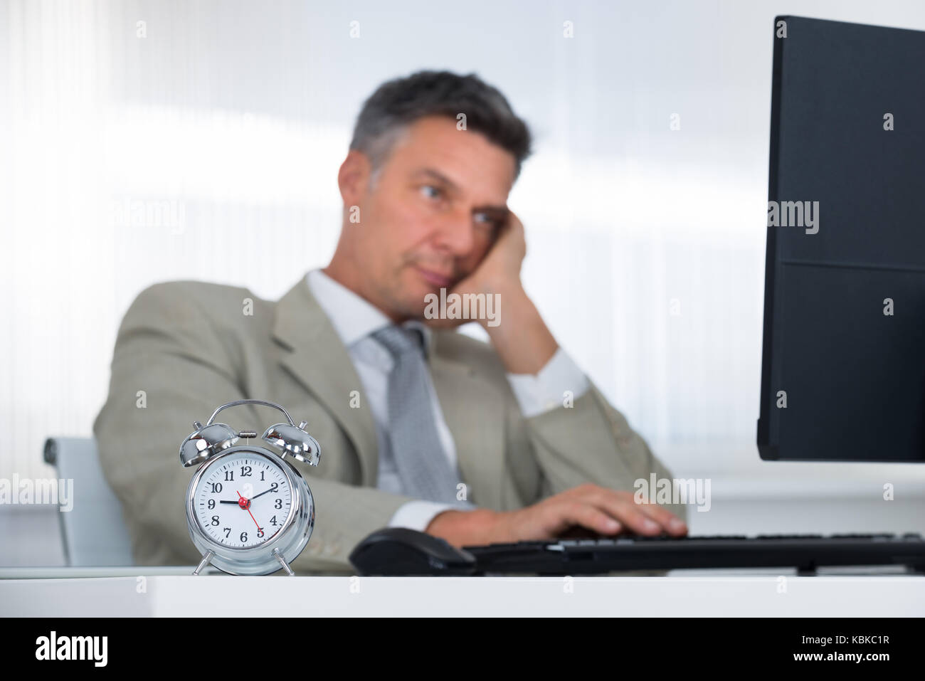 Tired businessman using computer at desk with focus on clock in office ...