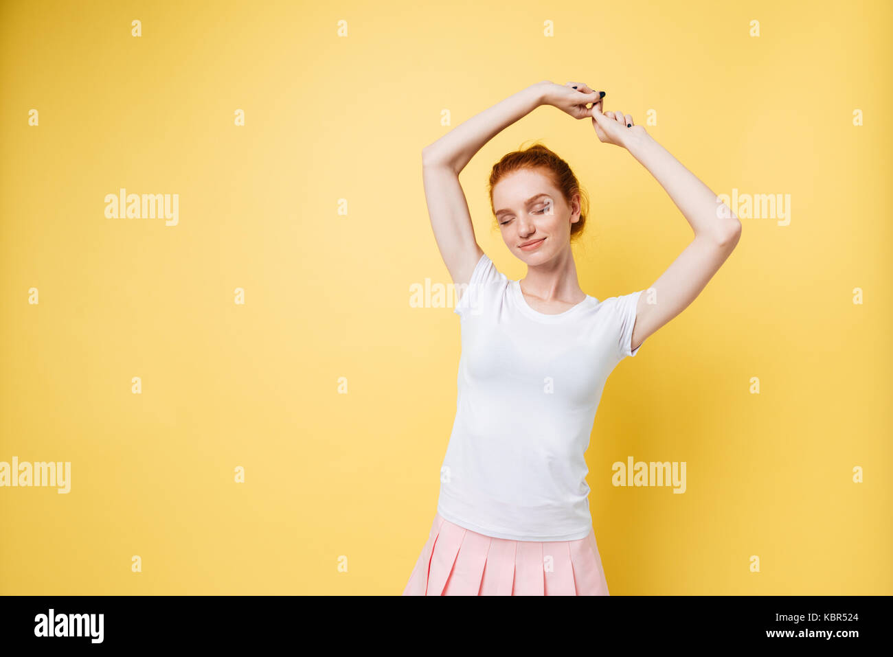 Pleased ginger girl posing with hand overhead and closed eyes over ...