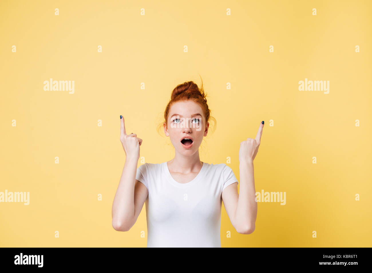 Shocked ginger woman pointing and looking up over yellow background ...