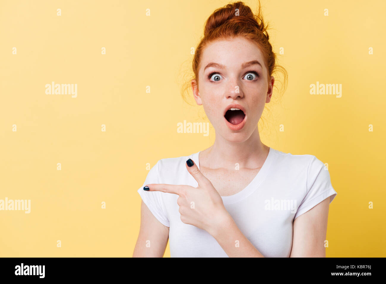 Shocked ginger woman in t-shirt pointing away and looking at the camera ...