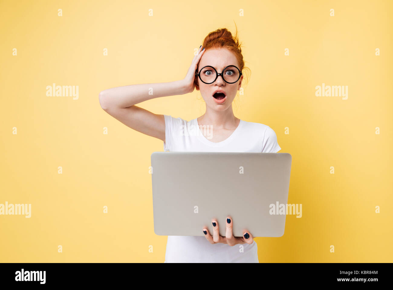 Shocked ginger woman in eyeglasses holding laptop computer in hand and ...