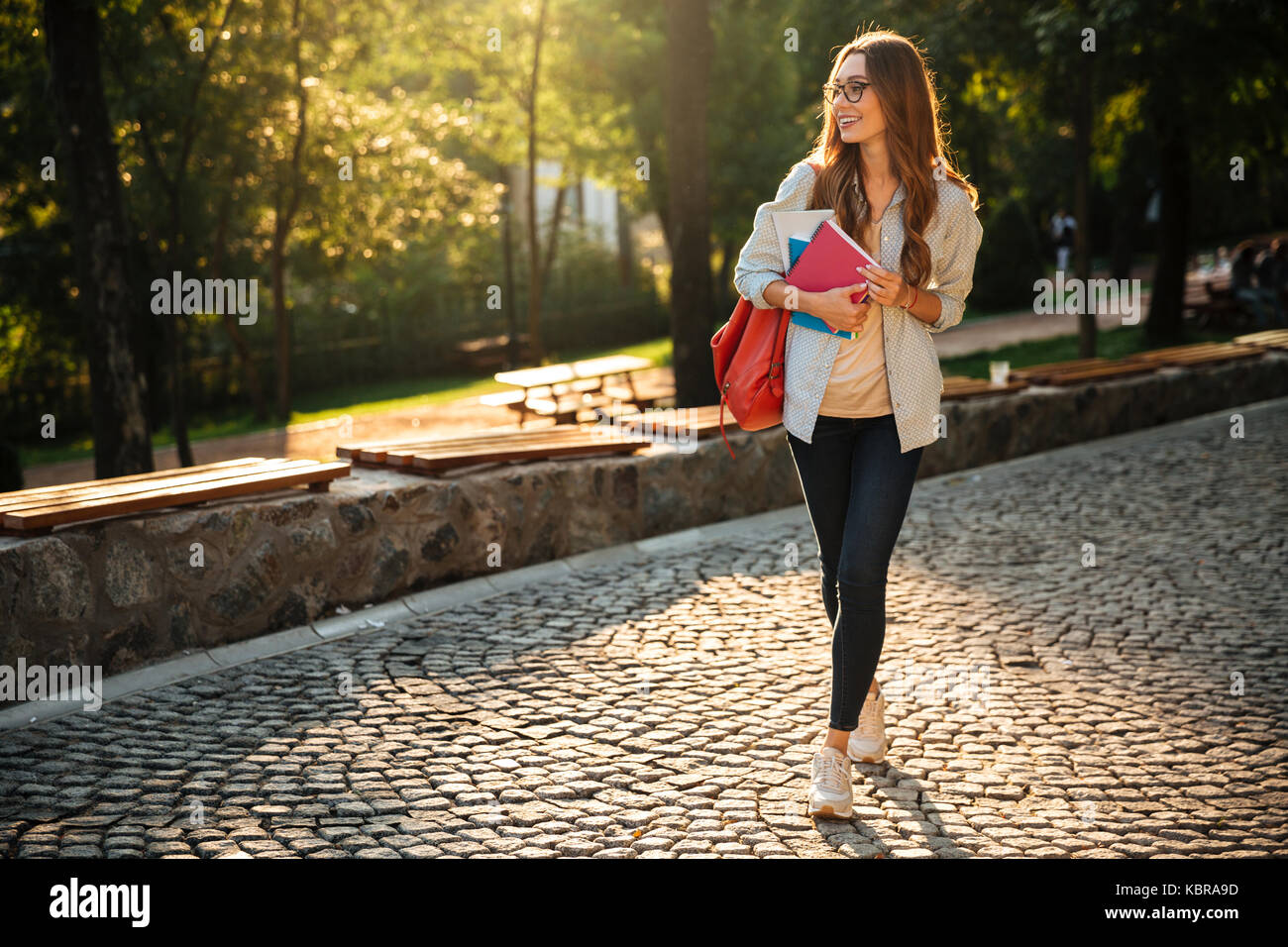Full length image of happy brunette woman walking with books in park ...