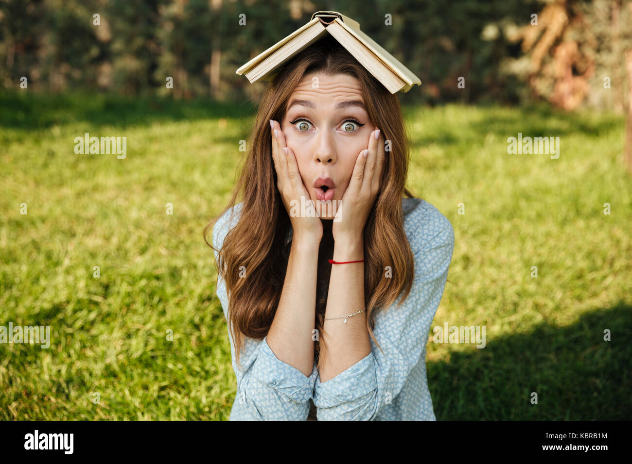Shocked brunette woman sitting in park with book on head while holding ...