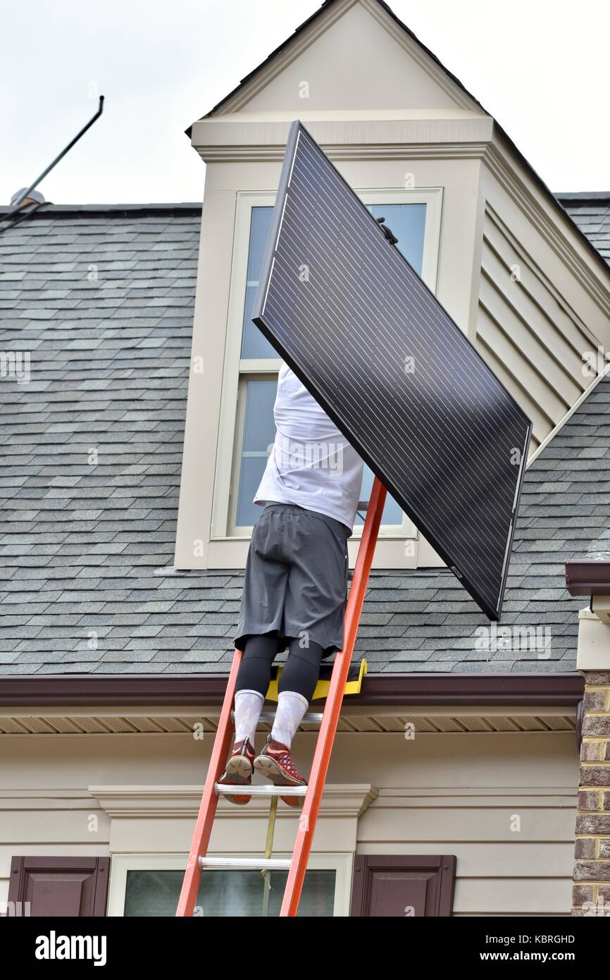 Solar panel technician carrying solar panels up ladder onto the roof of ...