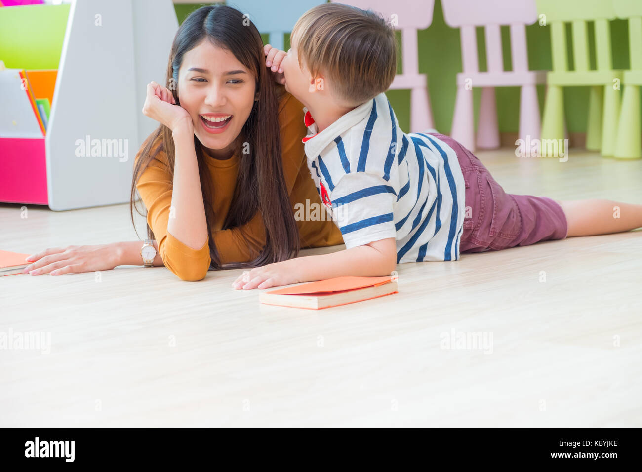 kid boy whisper teacher while lay down on library floor at kindergarten ...