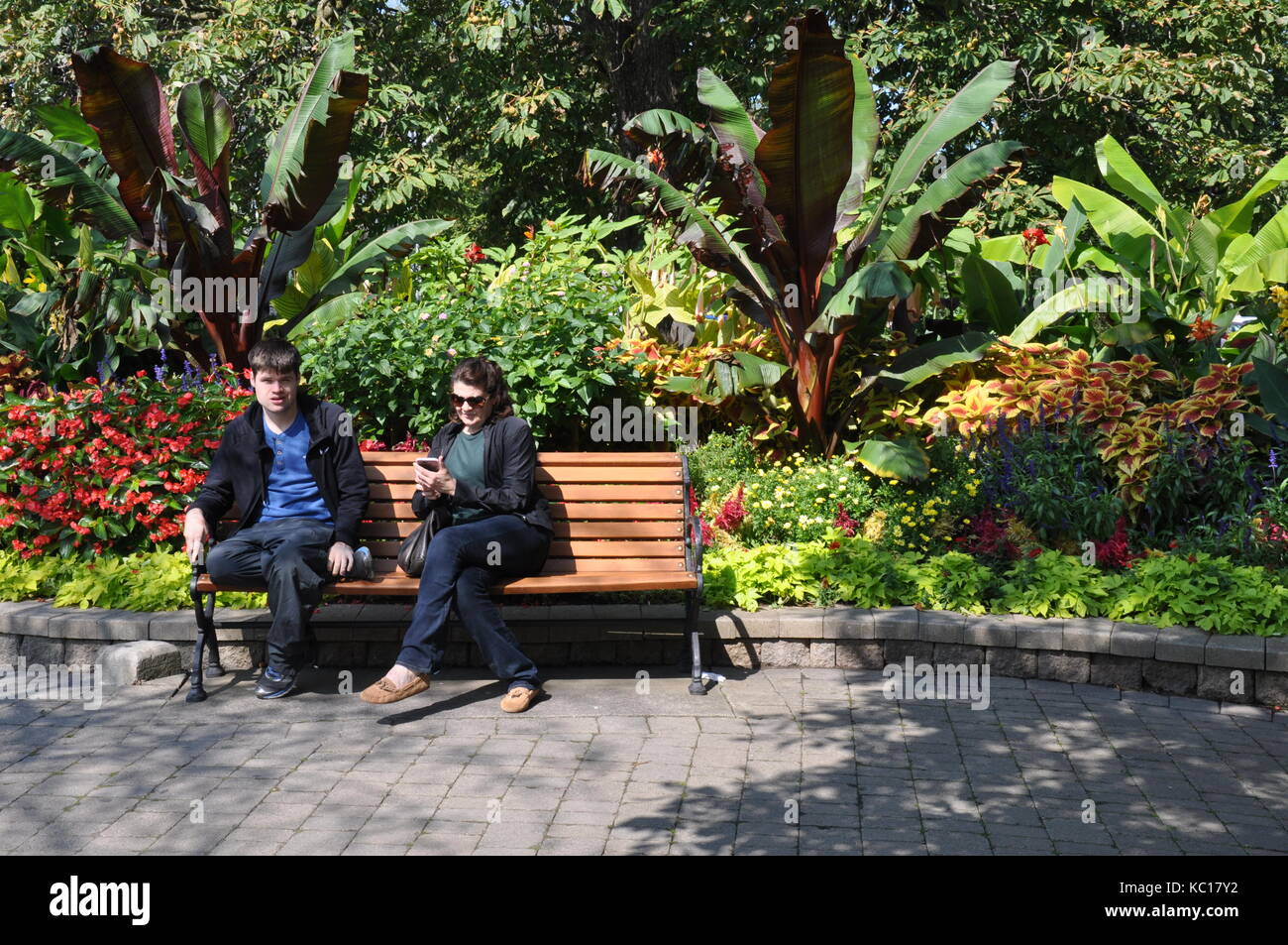 Two People Seated on a Bench in a City Park with Gardens in background ...