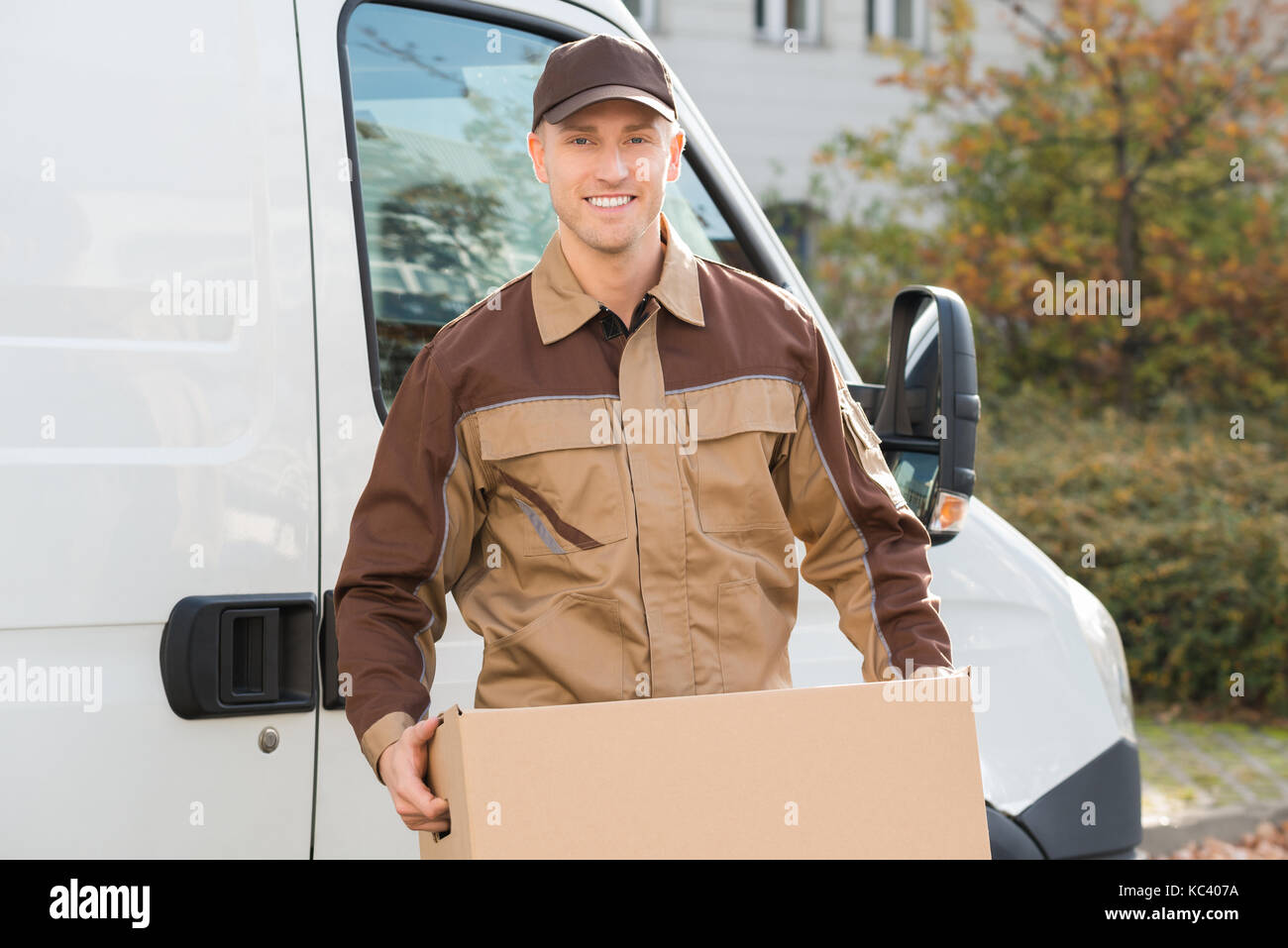 Portrait of young delivery man carrying cardboard box with truck in ...