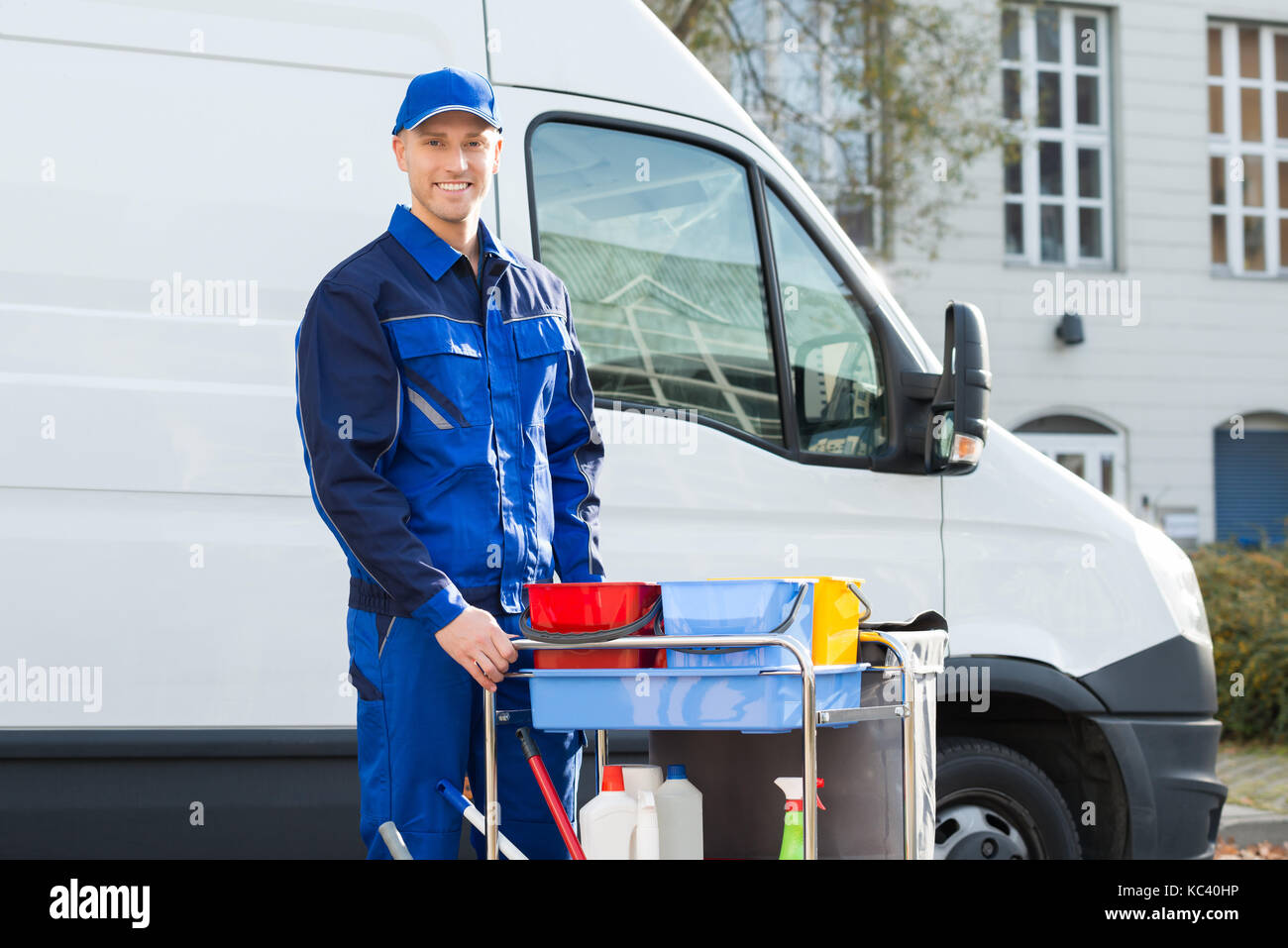 Portrait of happy male janitor standing with cleaning equipment against ...