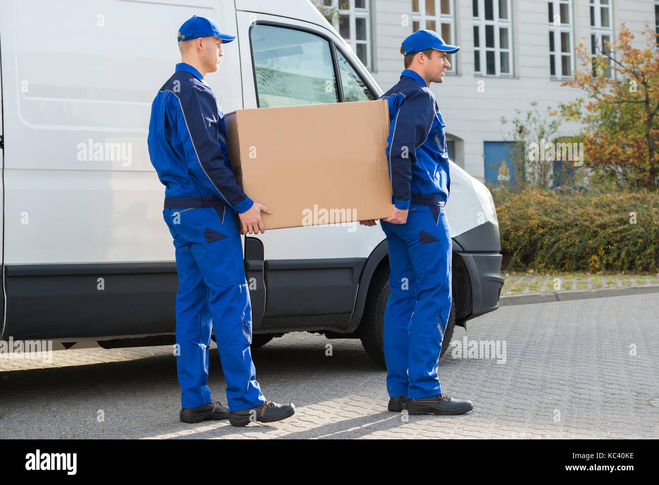 Side view of young delivery men carrying cardboard box while walking by ...