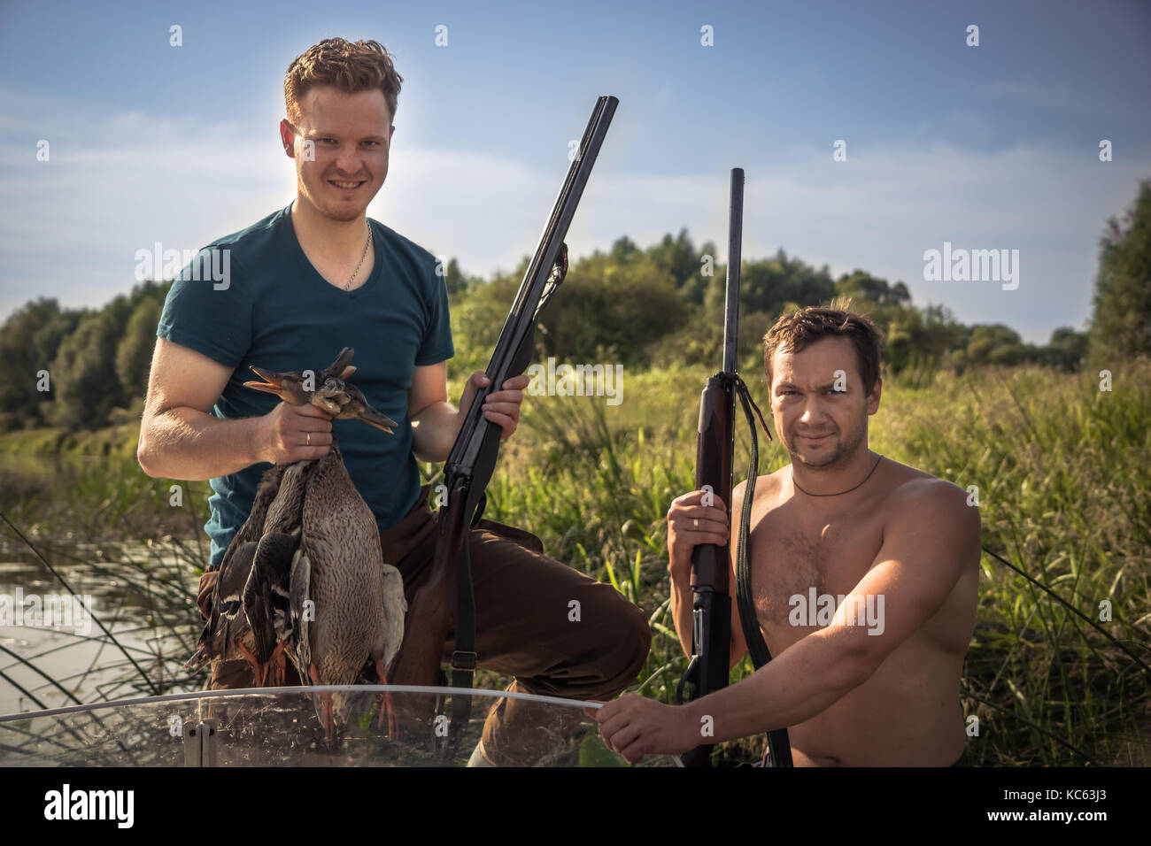 Cheerful men hunters with guns and trophy in boat on river bank during ...