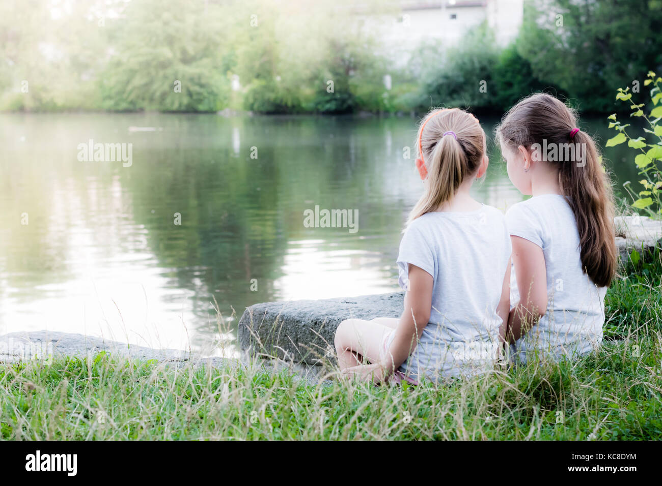 Two beautiful young girls are sitting near pond and looking to the ...