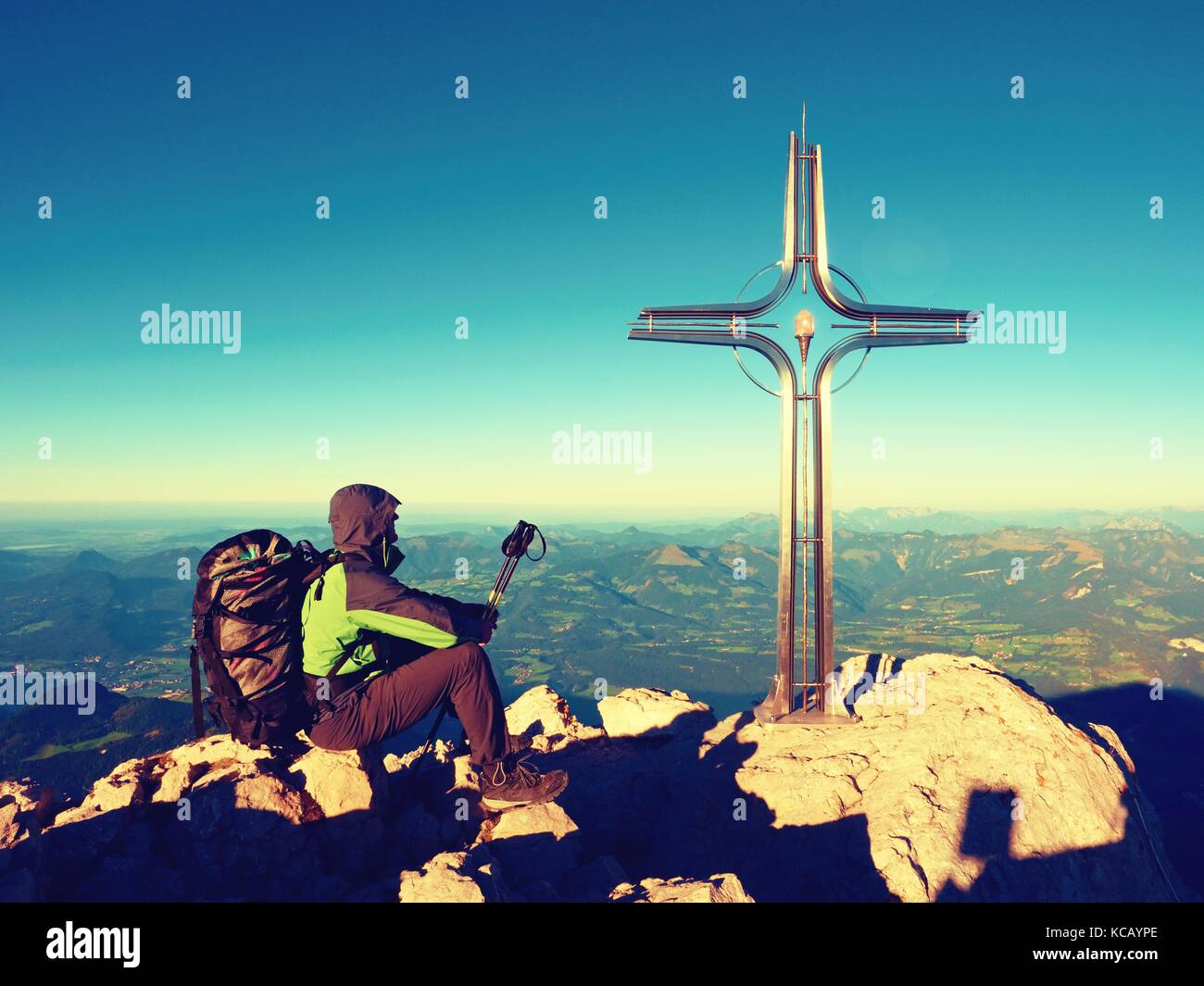 Tired hiker sit bellow crucifix on mountain peak. Iron cross at Alps ...