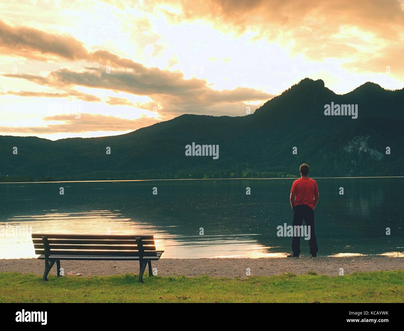 Tall man in red t-shirt at wooden bench at mountains lake coast. Dark ...