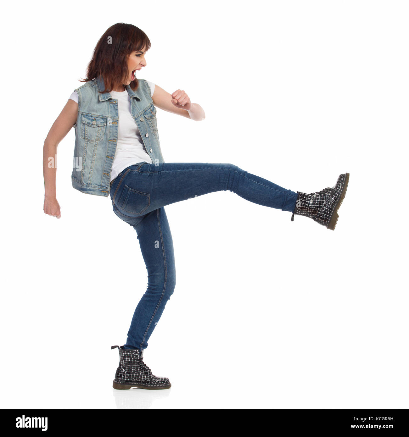 Young woman in jeans vest and black boots is shouting and kicking. Side ...
