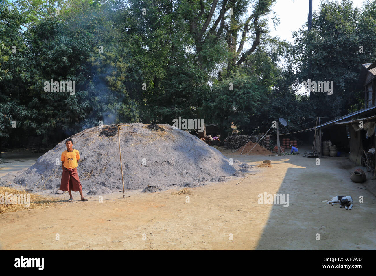 A man wearing a longyi stands beside a smoldering pile where clay pots ...