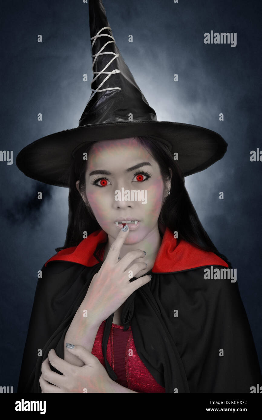 Vampire in witch suit with red blood on her lips in front of dark ...