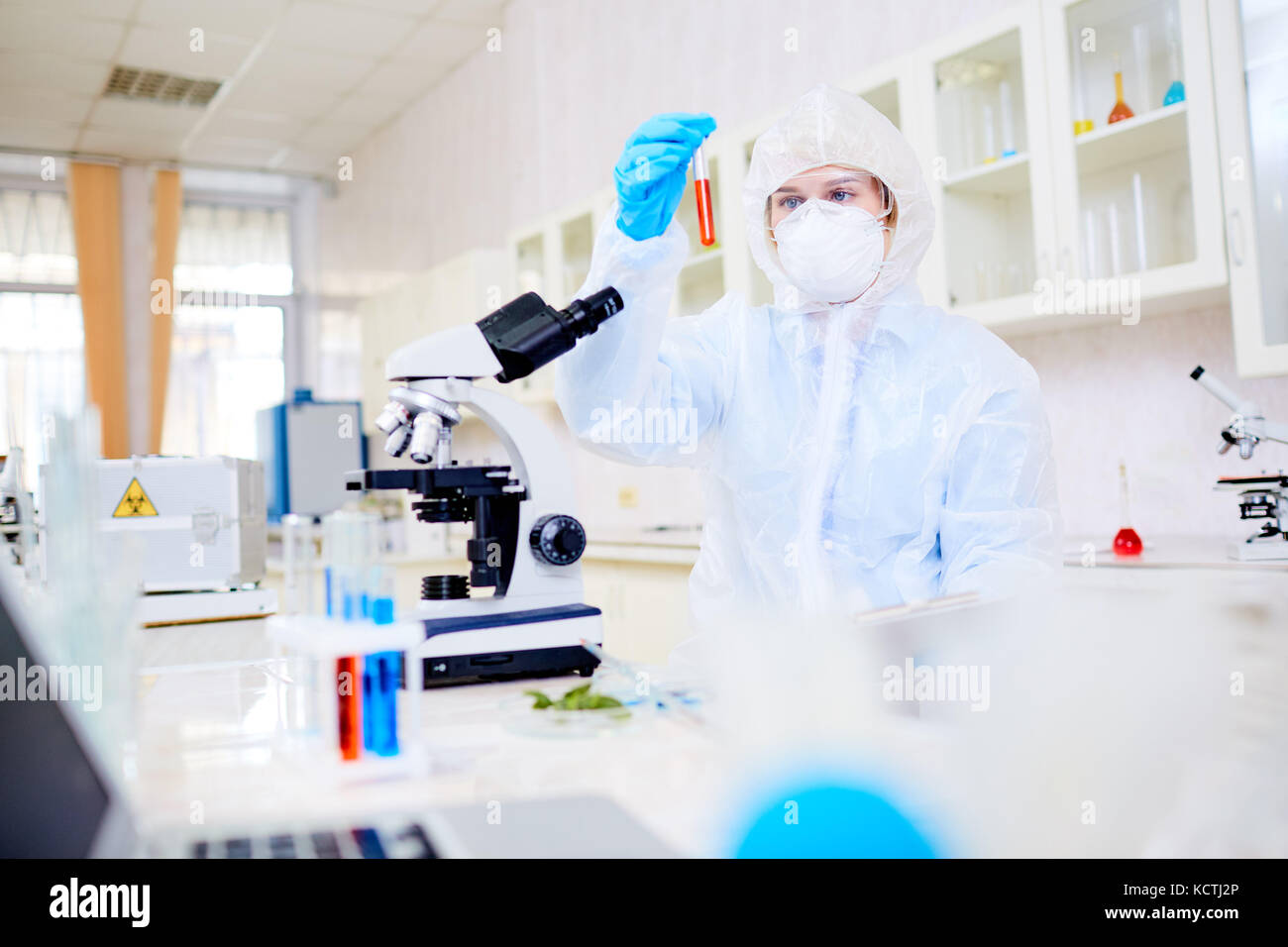 Female laboratory worker wearing filtering mask and coverall looking at ...