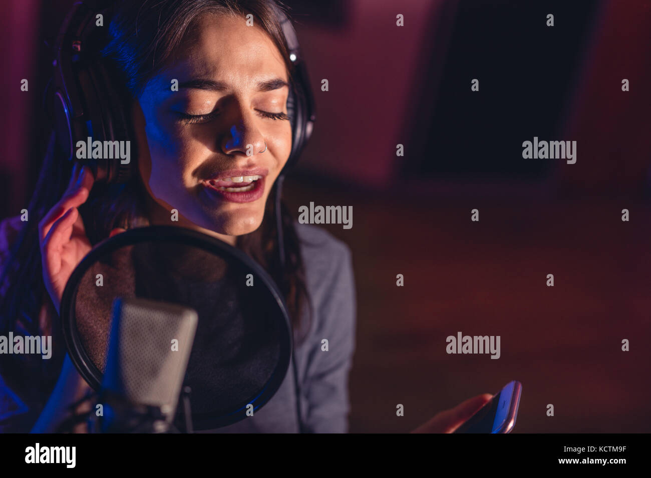 Young woman singing a song in the recording studio with mobile phone. Female singer in front of ...