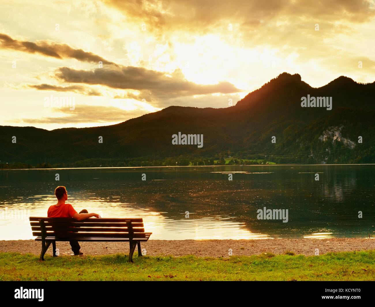 Alone man sits leg over on bench beside an azure mountain lake. Man ...