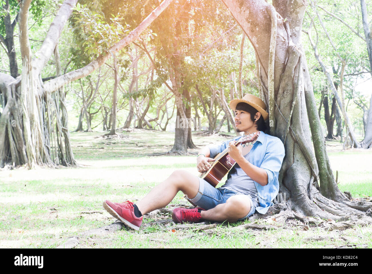 Happy asian man sitting under tree, wearing straw hat and playing ...