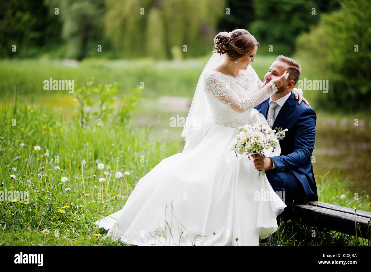 Fantastic bride sitting on a groom's lap in the meadow next to the lake ...