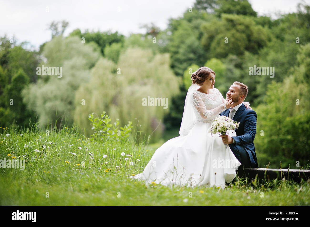 Fantastic bride sitting on a groom's lap in the meadow next to the lake ...