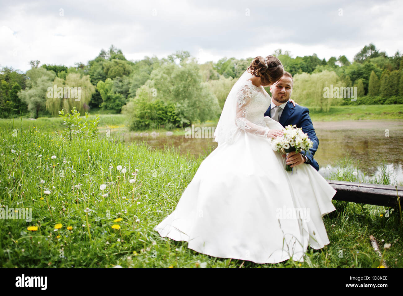 Fantastic bride sitting on a groom's lap in the meadow next to the lake ...