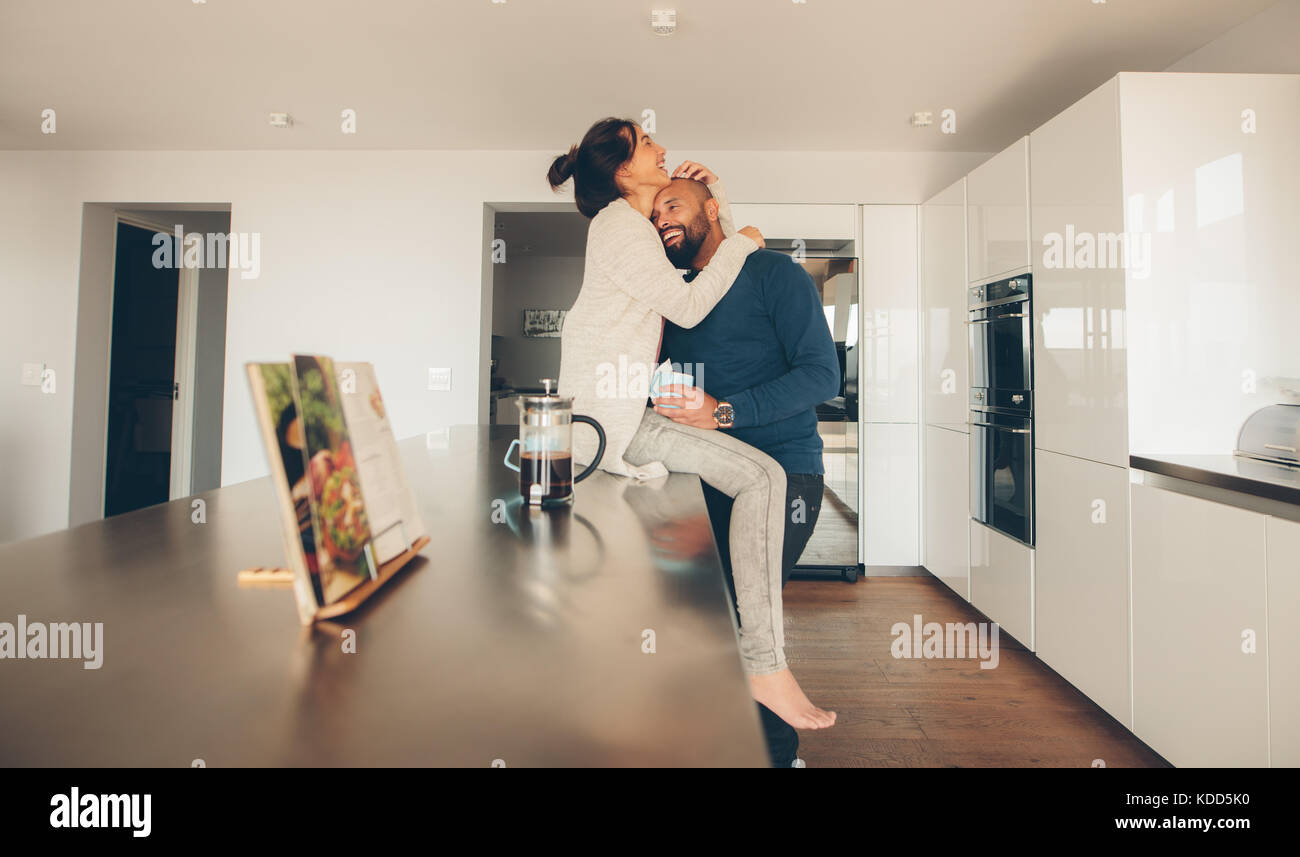 Happy young woman sitting on kitchen counter and embracing her man ...