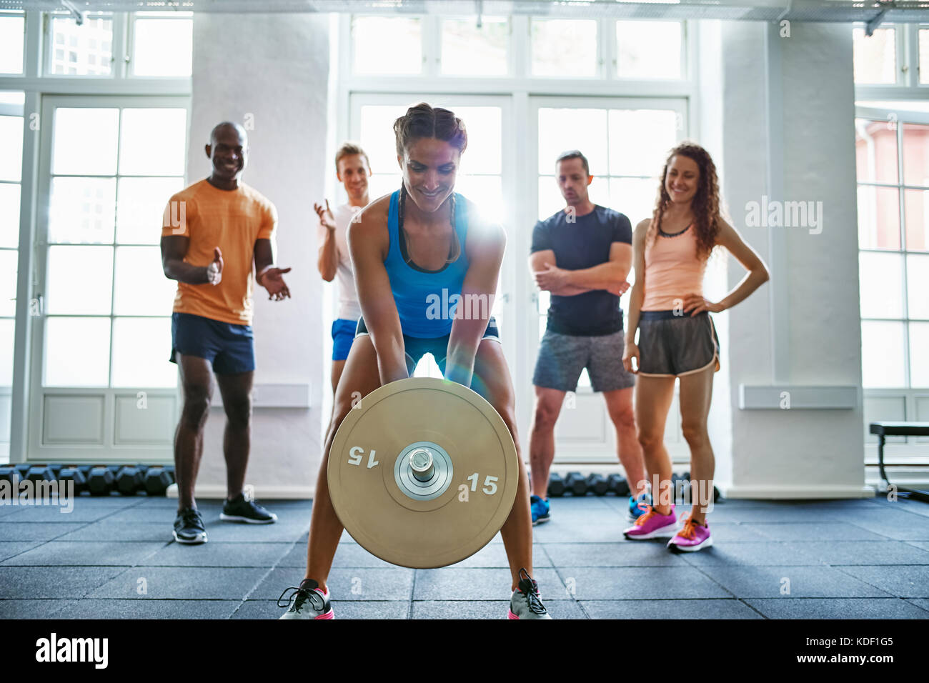 Group of young people cheering on their female friend exercising with weights while working out ...