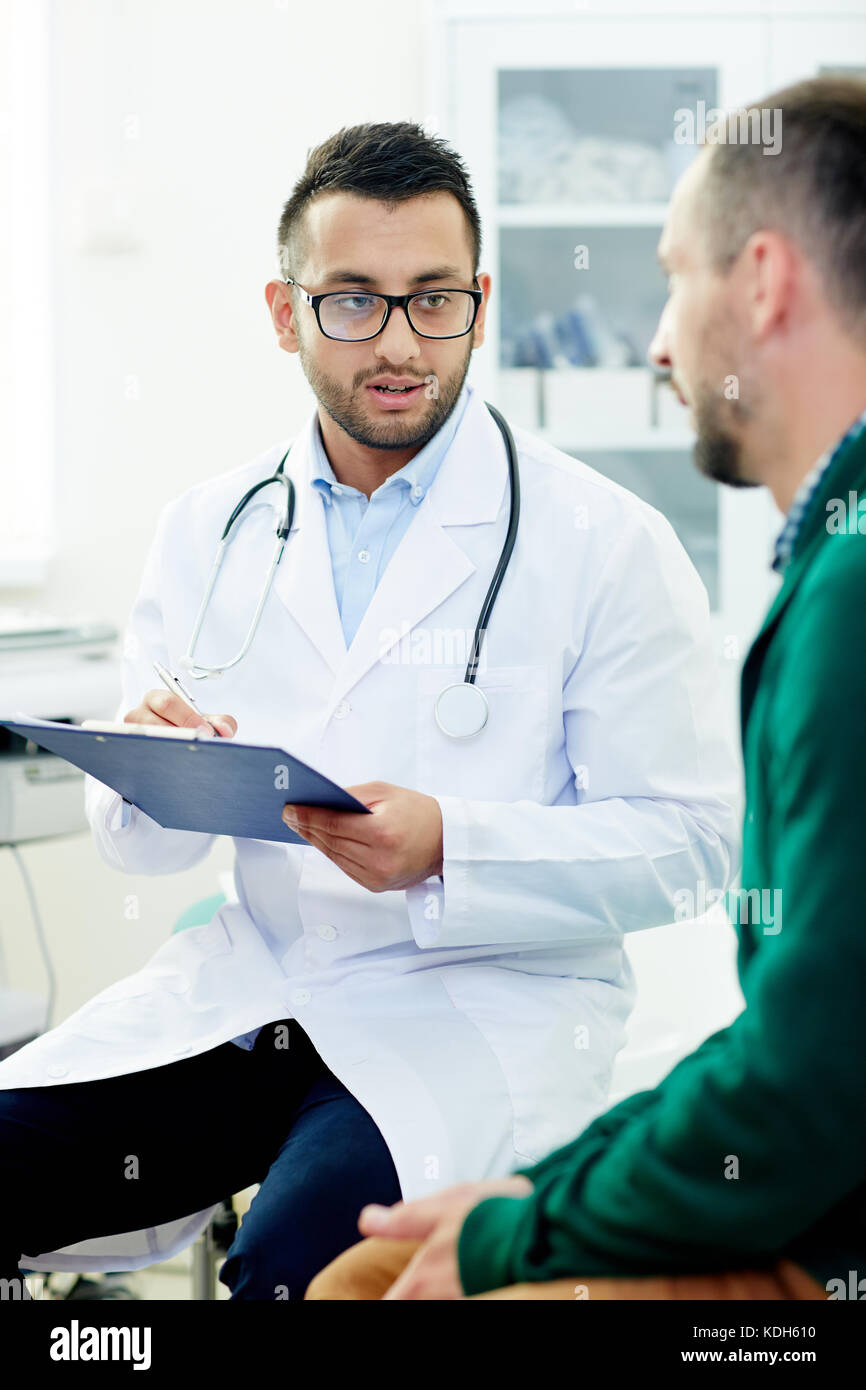 Young clinician making notes in medical card of patient while talking ...