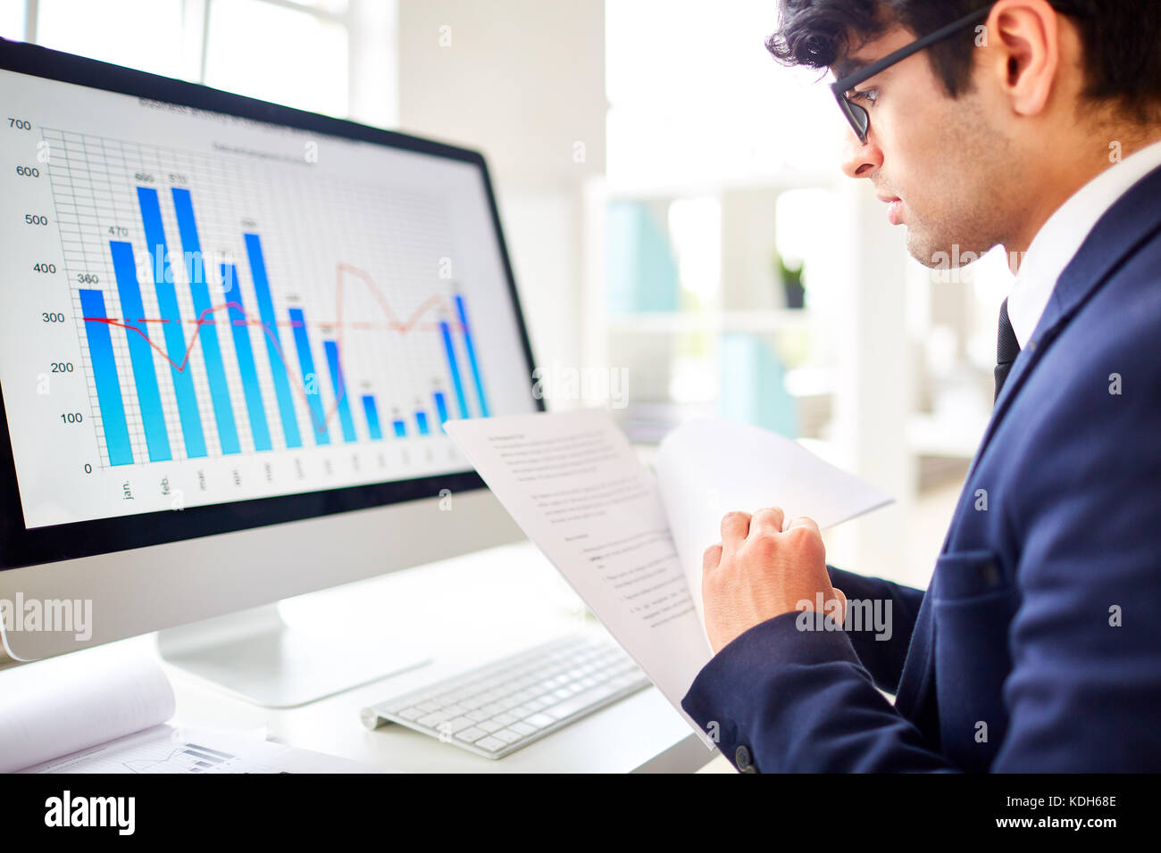 Modern economist looking through papers in front of computer monitor ...
