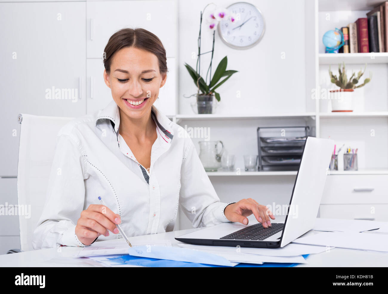cheerful woman writing down information from laptop at company office ...