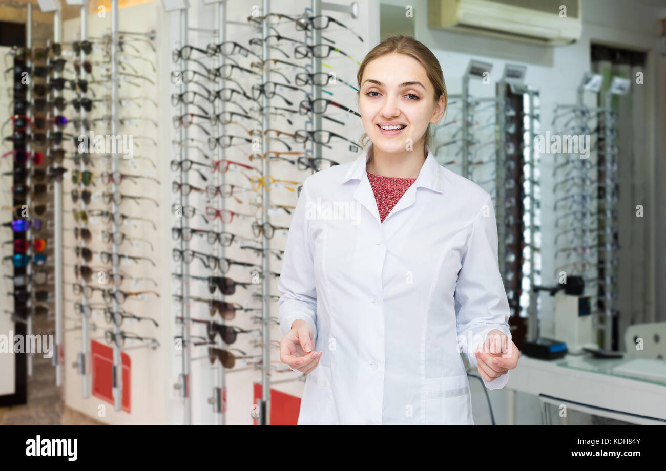 Professional female optician posing near stand with spectacles and ...