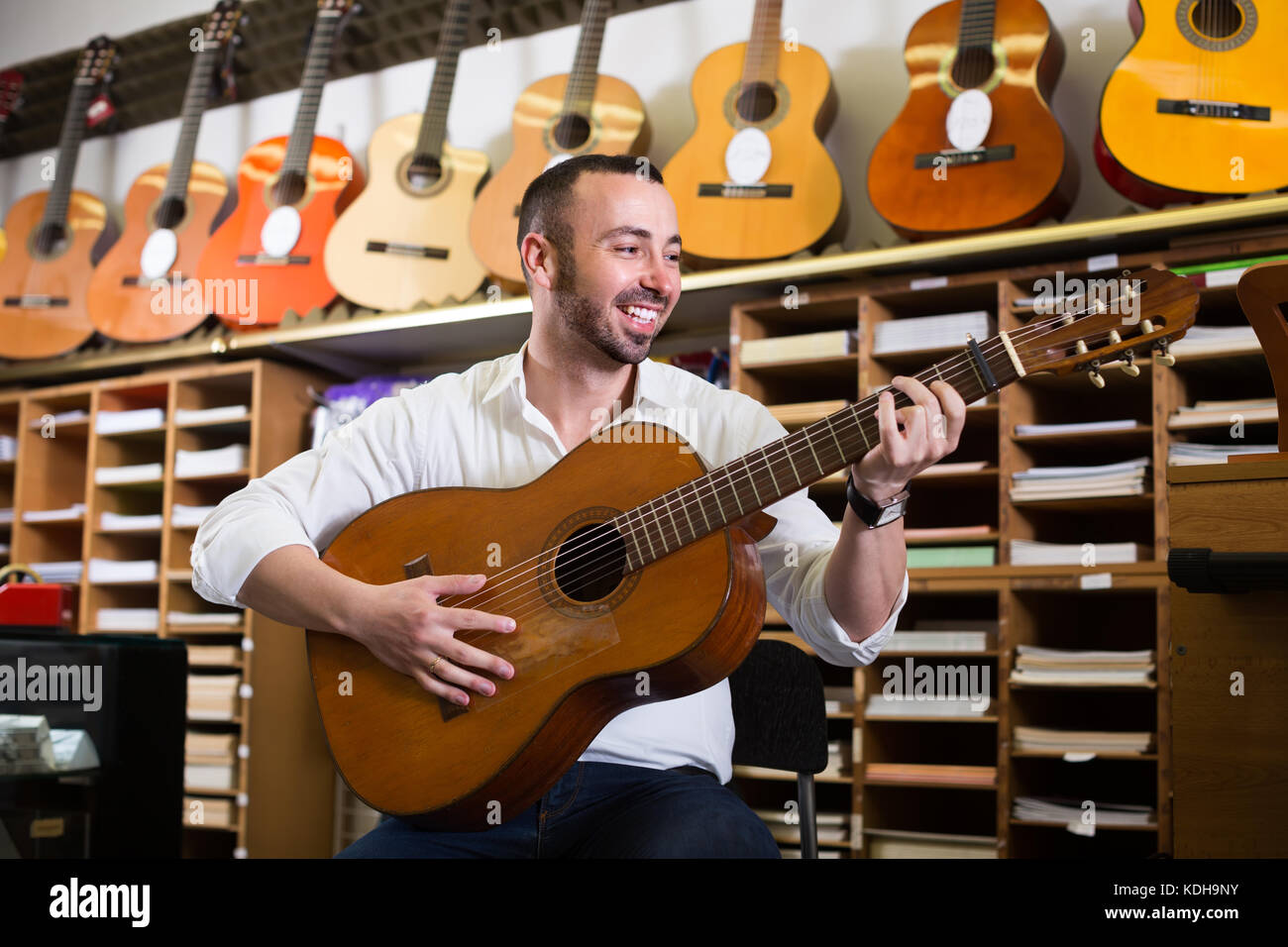 Joyful smiling young guy choosing acoustic guitar in music instruments ...