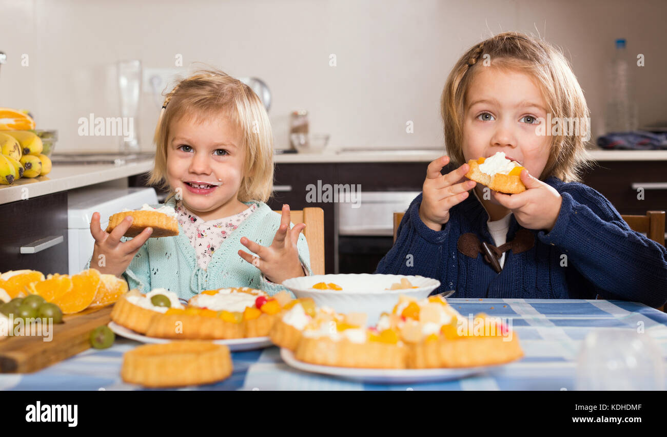 Two smiling little girls enjoying pastry with cream in kitchen at home ...