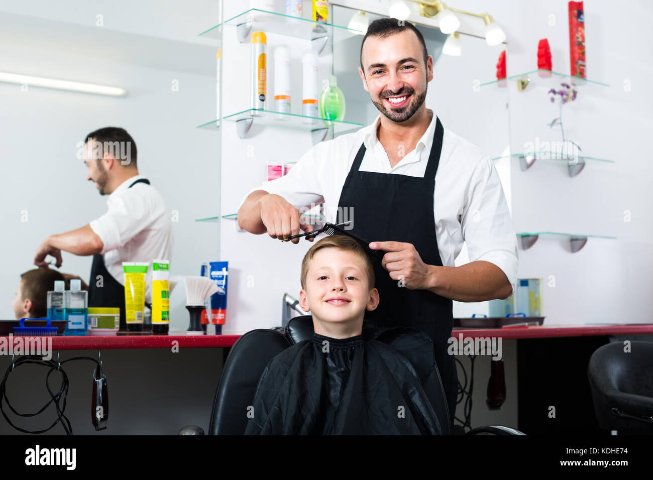 cheerful man hairdresser cutting hair of smiling boy in beauty salon ...