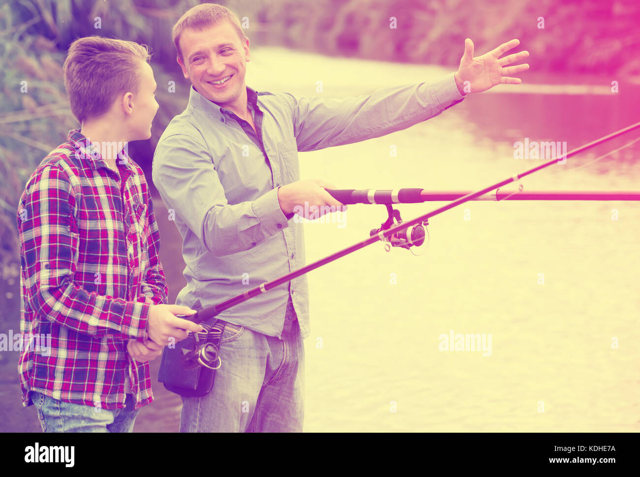 Smiling man and boy fishing together on freshwater lake from shore ...