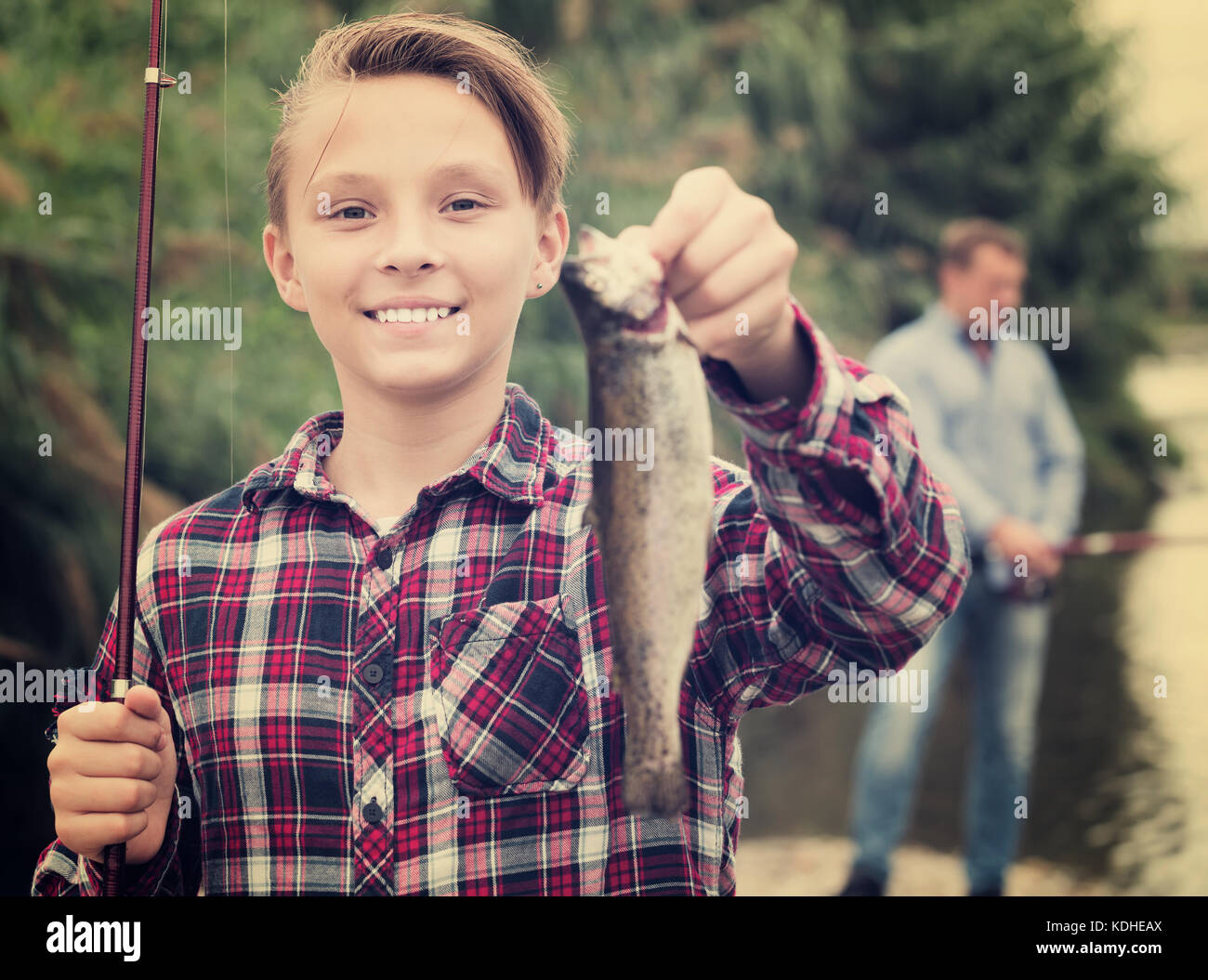 Cheerful teenage boy holding catch freshwater fish in hands on weekend ...