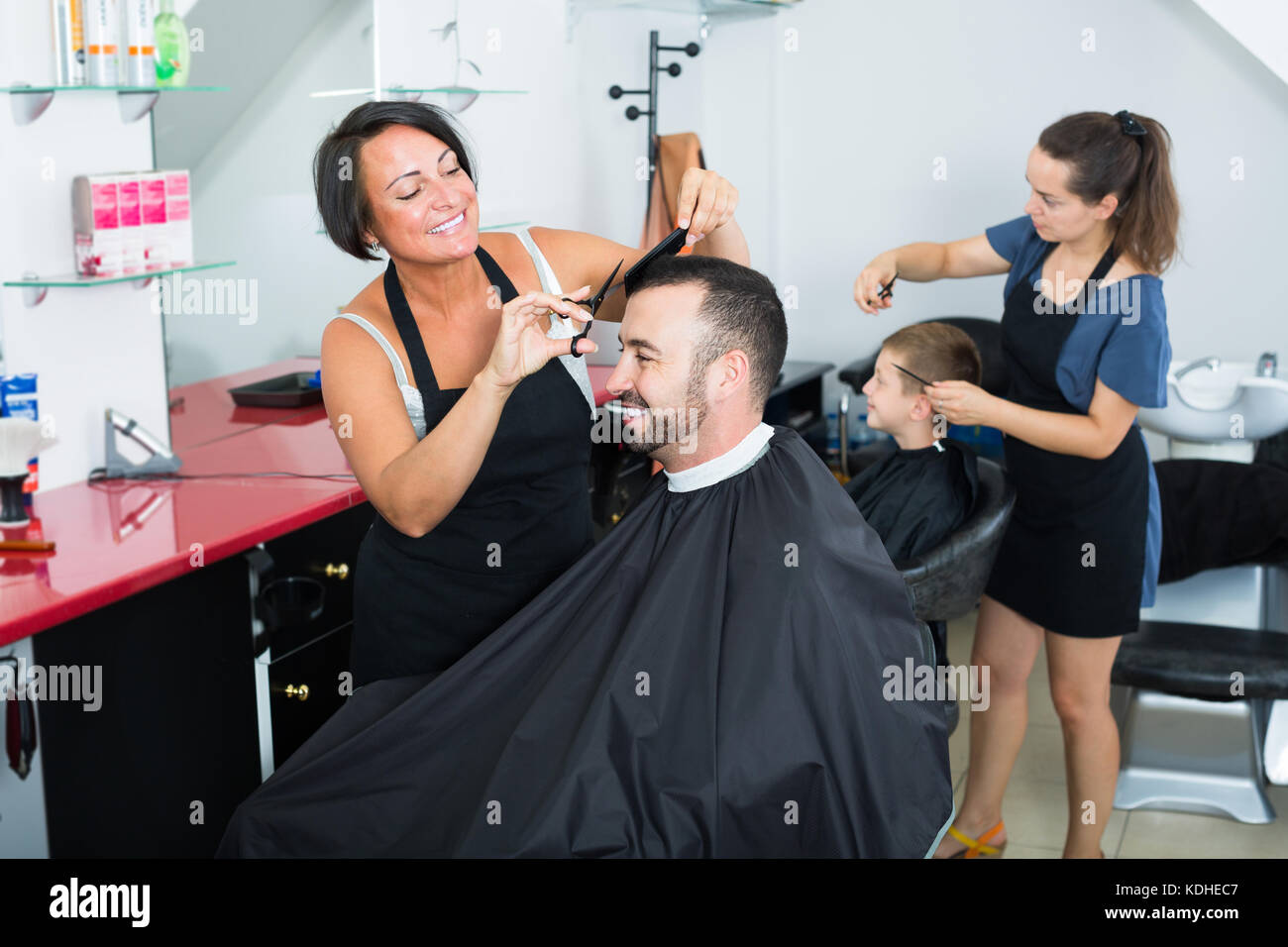Positive woman hairdresser with instrument cutting smiling male client ...