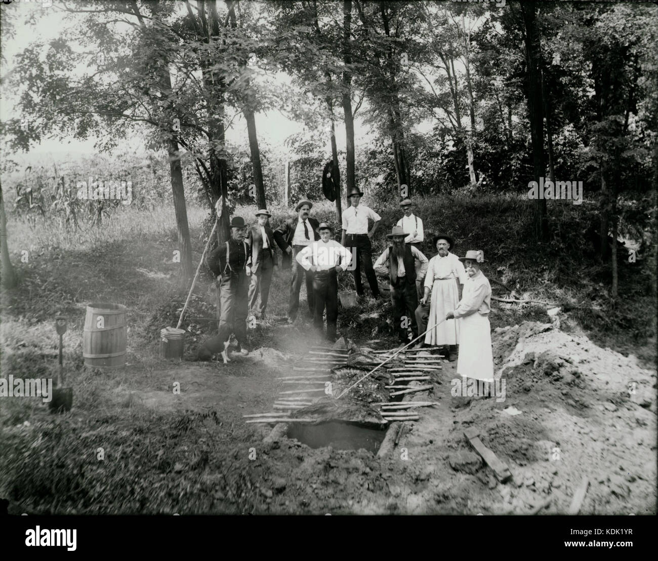 Nine men (with dog) cooking spitted meat on pit barbeque in rural ...