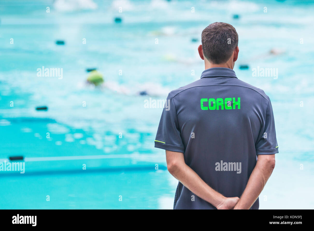 Male Swimming Coach Standing By The Swimming Pool Watching Swimmers