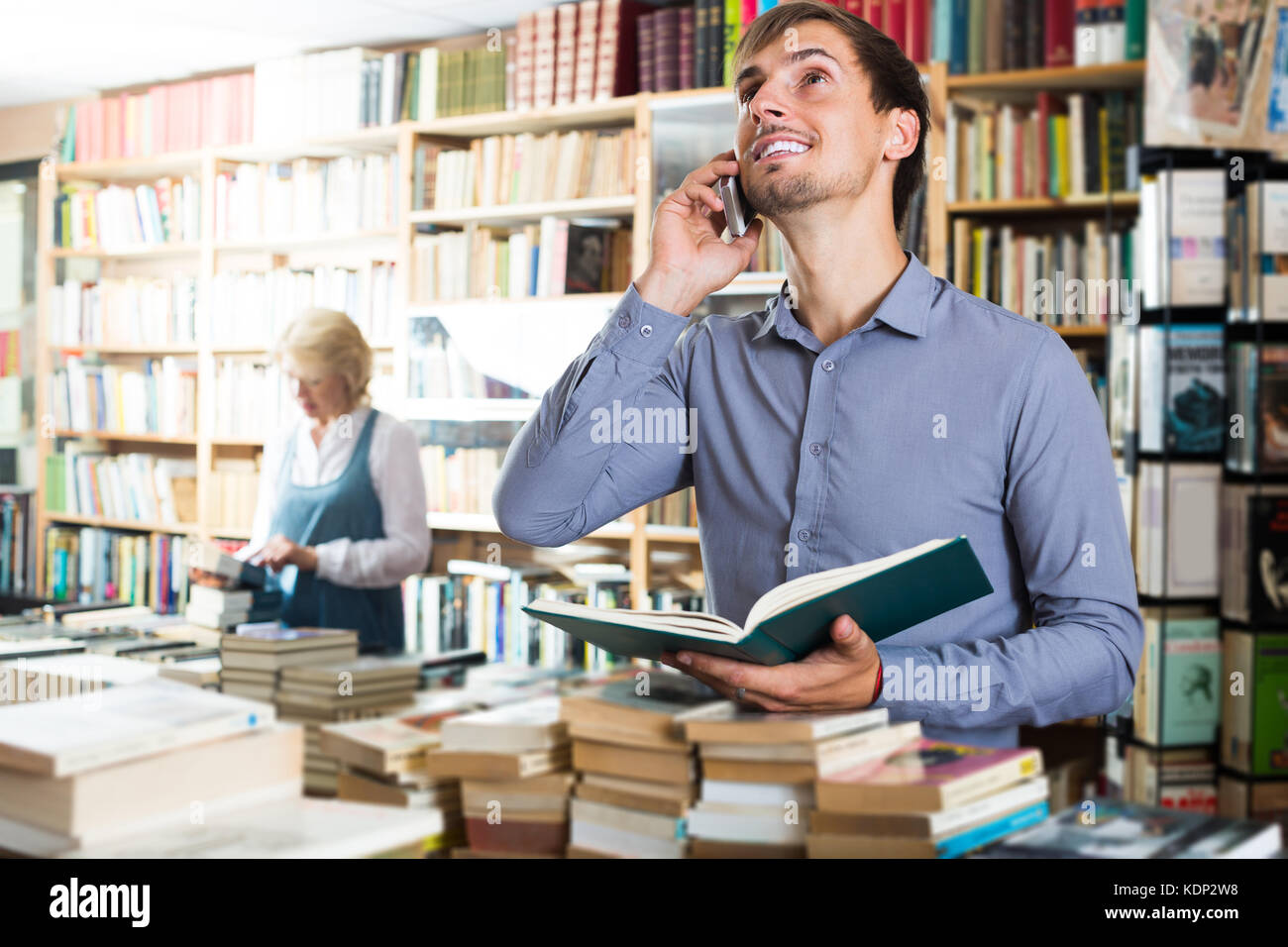 Young cheerful man looking at book and talking on mobile phone in book ...