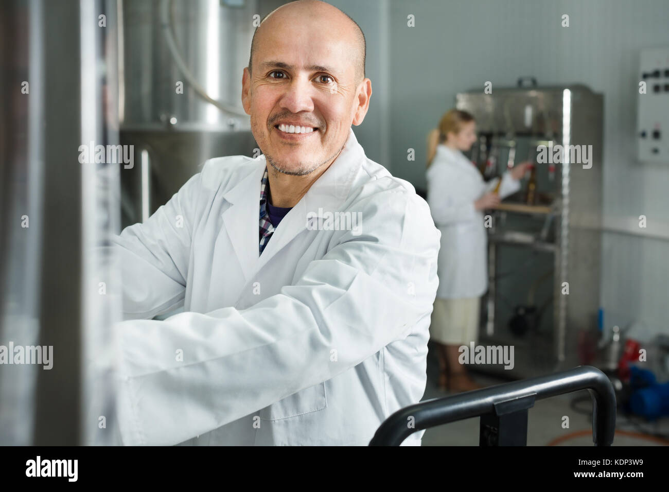 Mature cheerful man wearing uniform standing among a brewery stainless ...