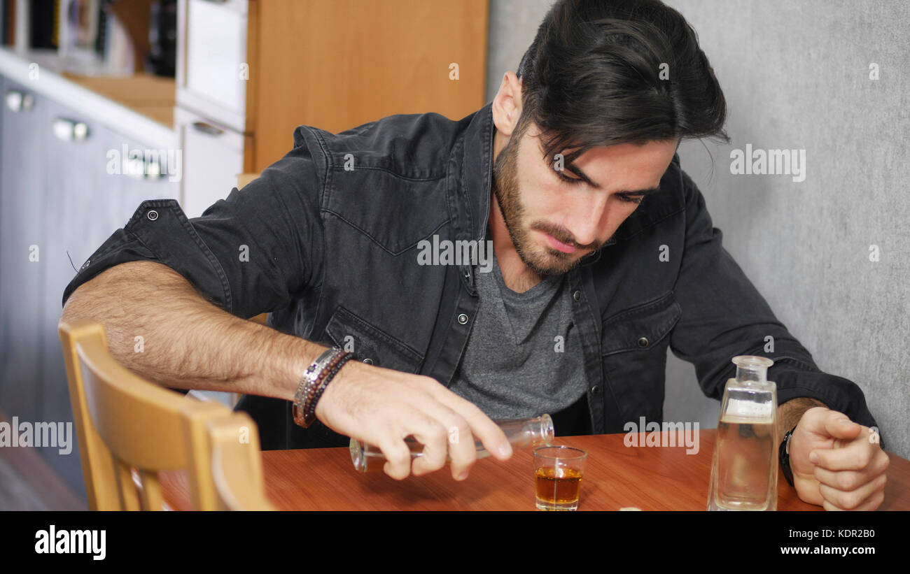 Young man sitting drinking alone at a table with two bottles of liquor ...