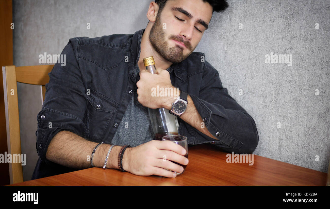 Young man sitting drinking alone at a table with two bottles of liquor ...