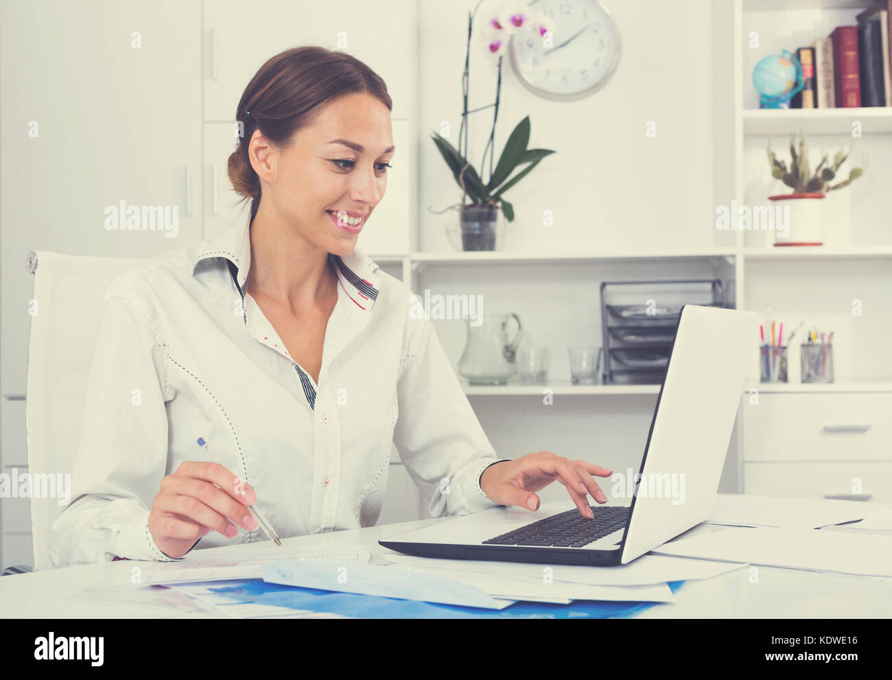 cheerful woman writing down information from laptop at company office ...